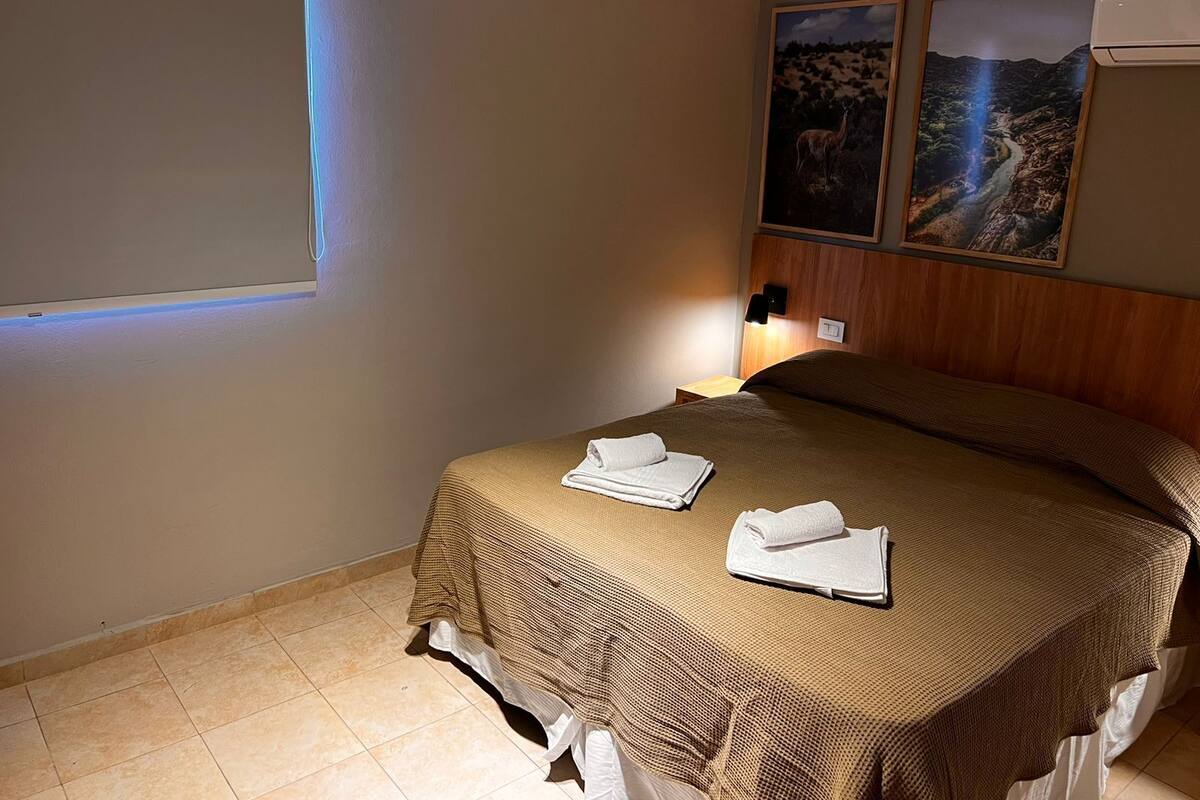 Quarto de hotel com cama, mesa de cabeceira, luminária na parede e duas fotos emolduradas.