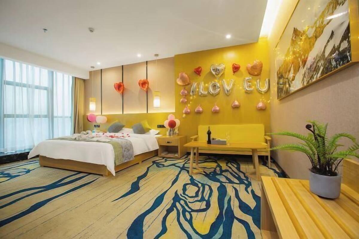 Quarto de hotel com cama, mesas de cabeceira, escrivaninha, uma cadeira, planta em vaso e uma janela grande.