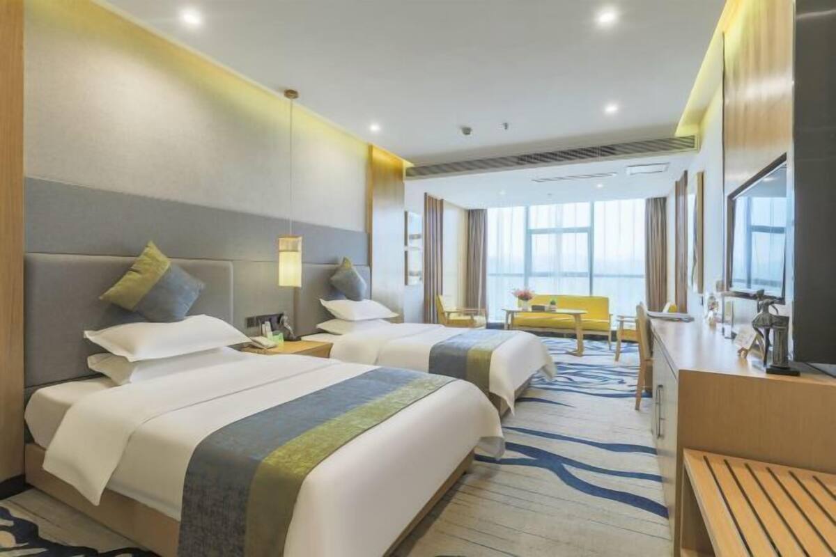 Quarto de hotel com duas camas, uma escrivaninha, uma cadeira e uma janela grande.