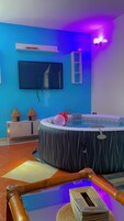 Quarto com uma grande banheira de hidromassagem redonda, televisão de tela plana na parede, um pequeno armário branco e uma prateleira com objetos decorativos.