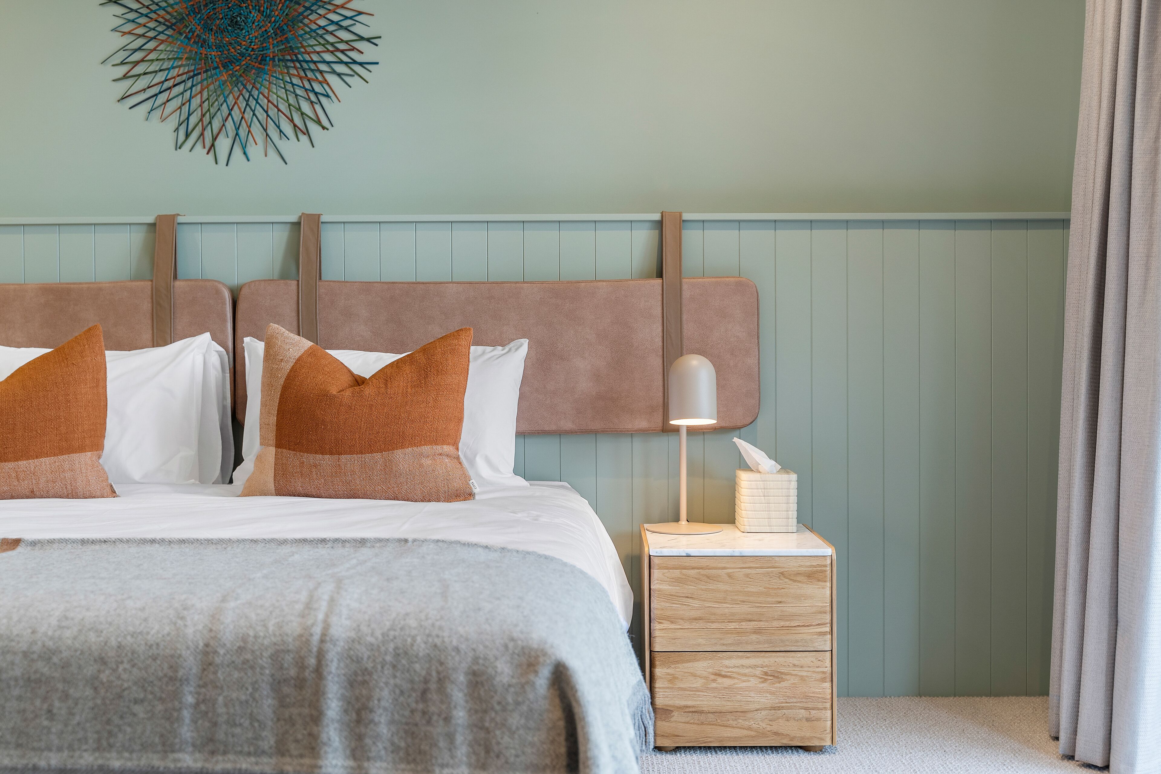 Cama de casal bem arrumada com manta cinza, duas almofadas castanhas e mesa de cabeceira de madeira com abajur e caixa de lenços.
