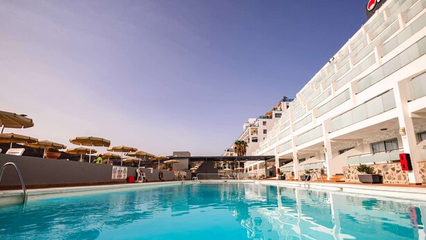 Piscina com guarda-sóis, um edifício moderno com várias varandas e céu claro.