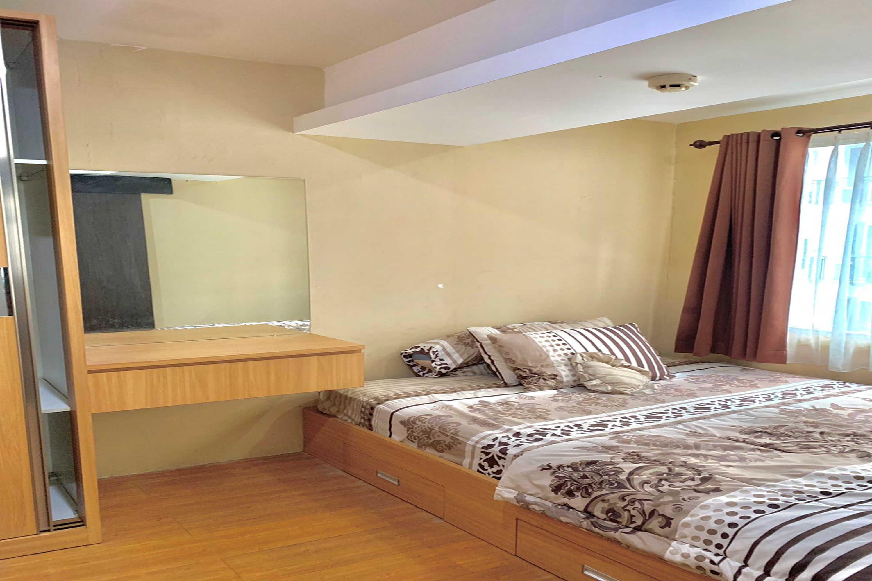 Quarto com cama, escrivaninha de madeira, espelho e janela com cortinas.