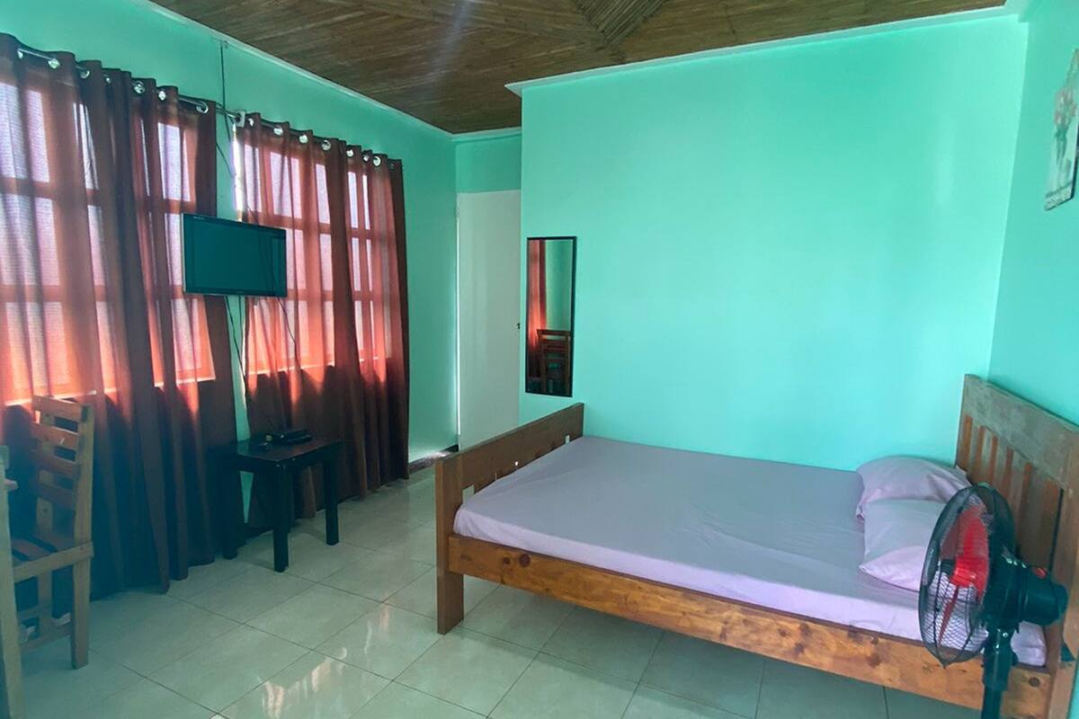 Quarto com teto de madeira, uma cama, escrivaninha com cadeira, ventoinha e televisão fixada na parede.