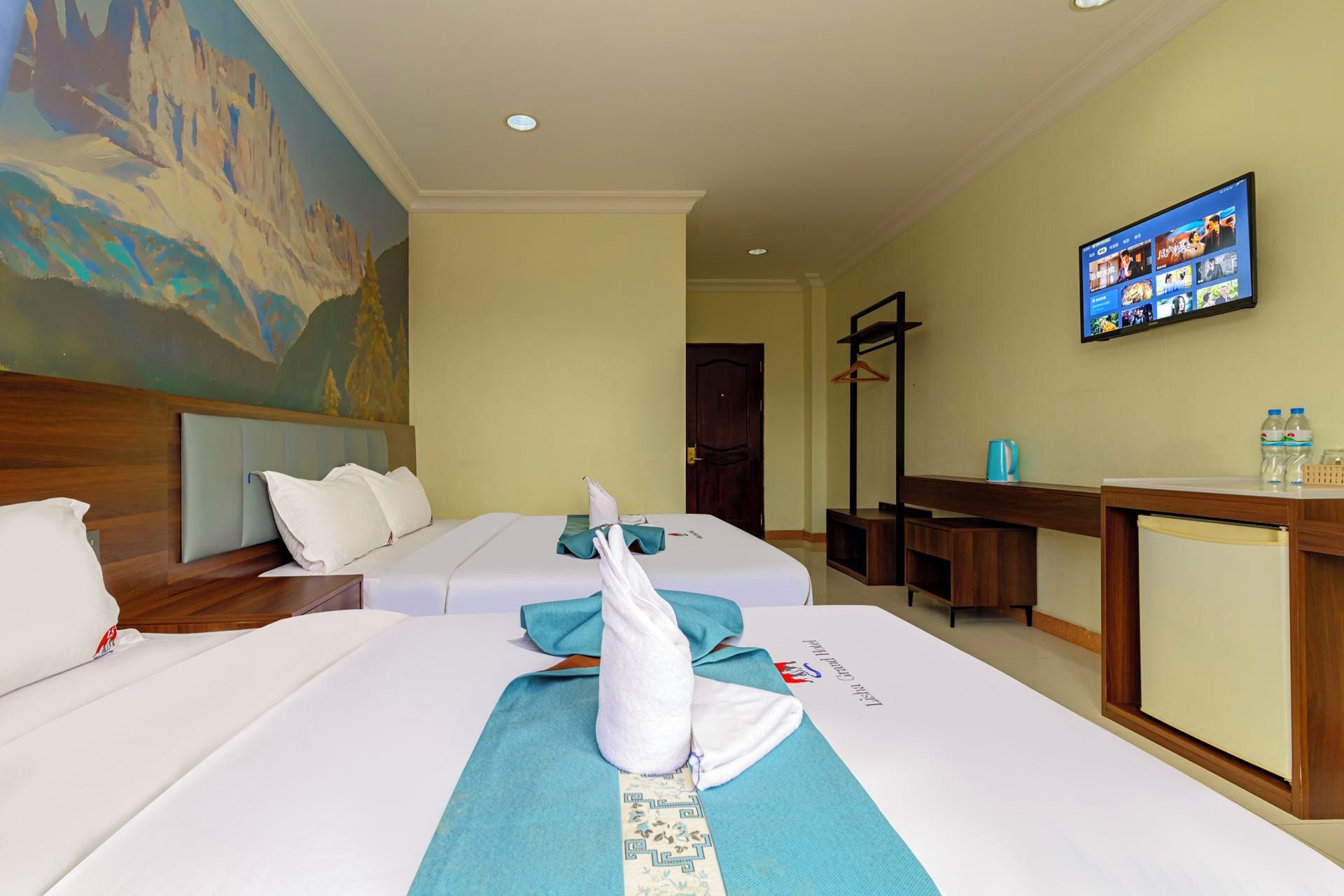 Quarto de hotel com duas camas, um quadro na parede, uma televisão e uma mesa com cafeteira e garrafas de água.