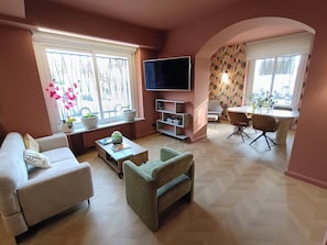 Uma sala de estar com sofá, poltronas, mesa de centro, televisão, área de jantar com mesa e cadeiras, e amplas janelas com plantas.