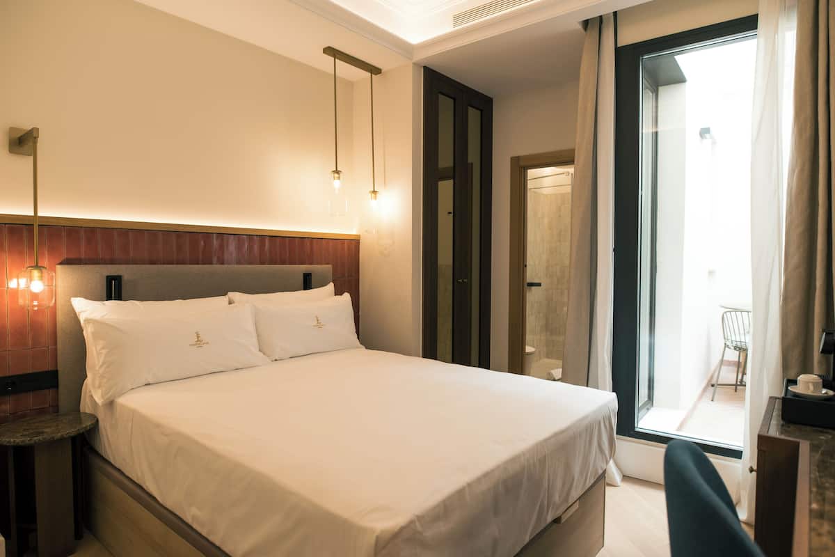 Quarto de hotel com uma cama grande, uma mesinha de cabeceira, uma cadeira e vista para a varanda externa.