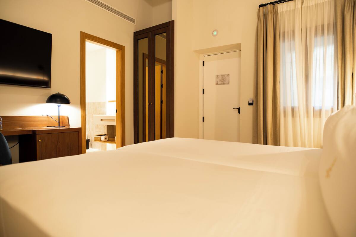 Quarto de hotel com uma cama grande, uma escrivaninha com abajur, uma TV na parede e uma porta com espelho.