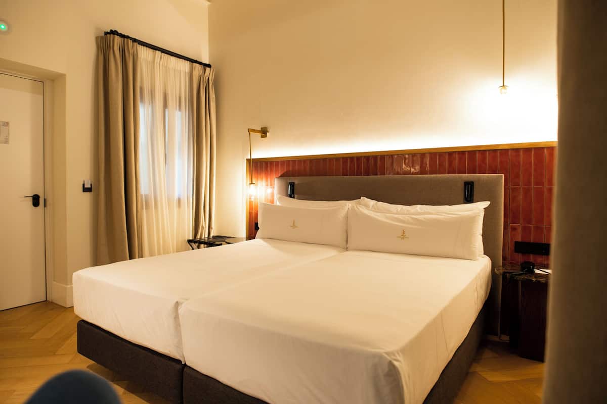 Quarto de hotel com uma cama grande, uma mesa de cabeceira, um abajur e uma cortina.
