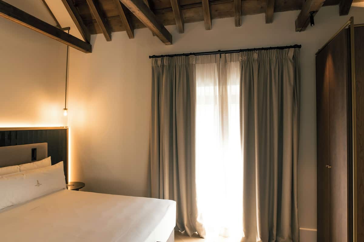 Quarto de hotel com cama, mesa de cabeceira, luminária e janela com cortinas.