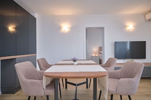 Uma sala de jantar moderna com uma mesa de madeira e seis cadeiras estofadas. Há uma pequena planta em vaso sobre a mesa, e o ambiente tem um design minimalista com uma televisão de tela plana na parede.