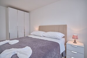 Cama bem arrumada com colcha cinza, travesseiros brancos e mesa de cabeceira com abajur.