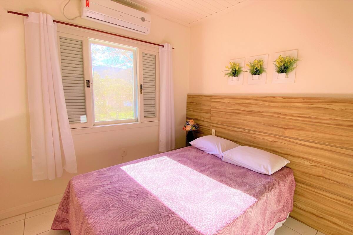 Quarto com cama, janela com persianas, ar condicionado e três plantas em vasos na parede.