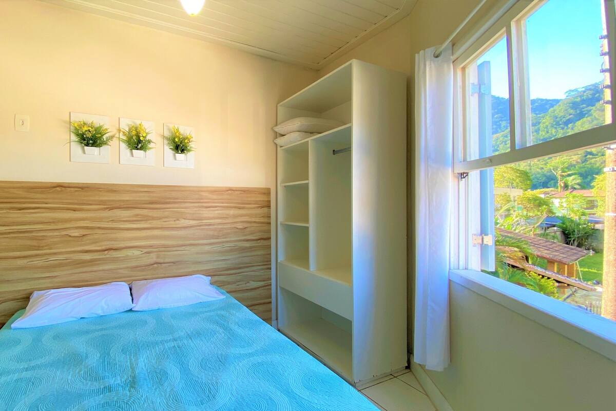 Quarto com cama, cabeceira de madeira, janela com vista e três plantas em vasos na parede.