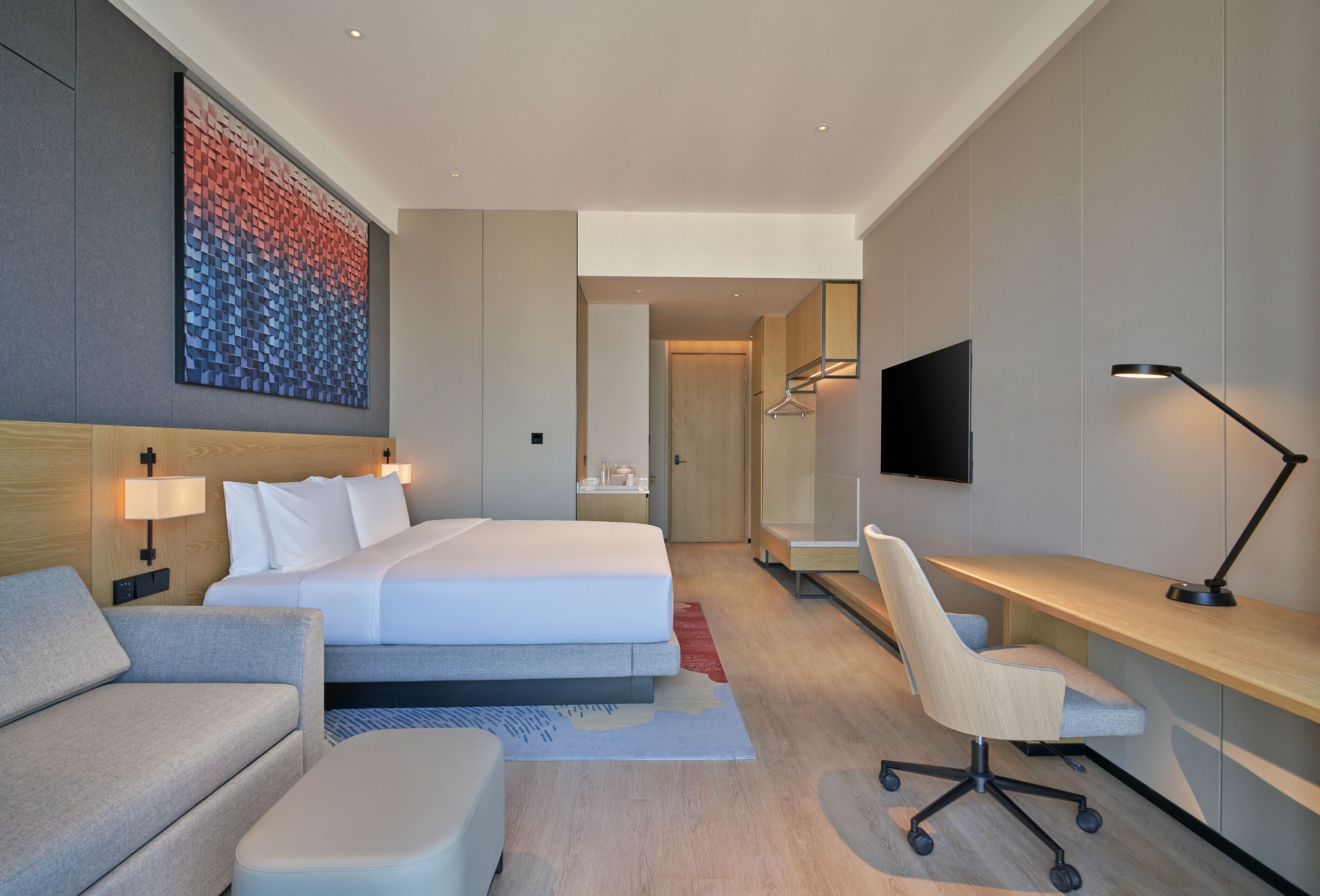 Quarto de hotel moderno com uma cama grande, uma escrivaninha, uma cadeira, uma televisão e um quadro na parede.
