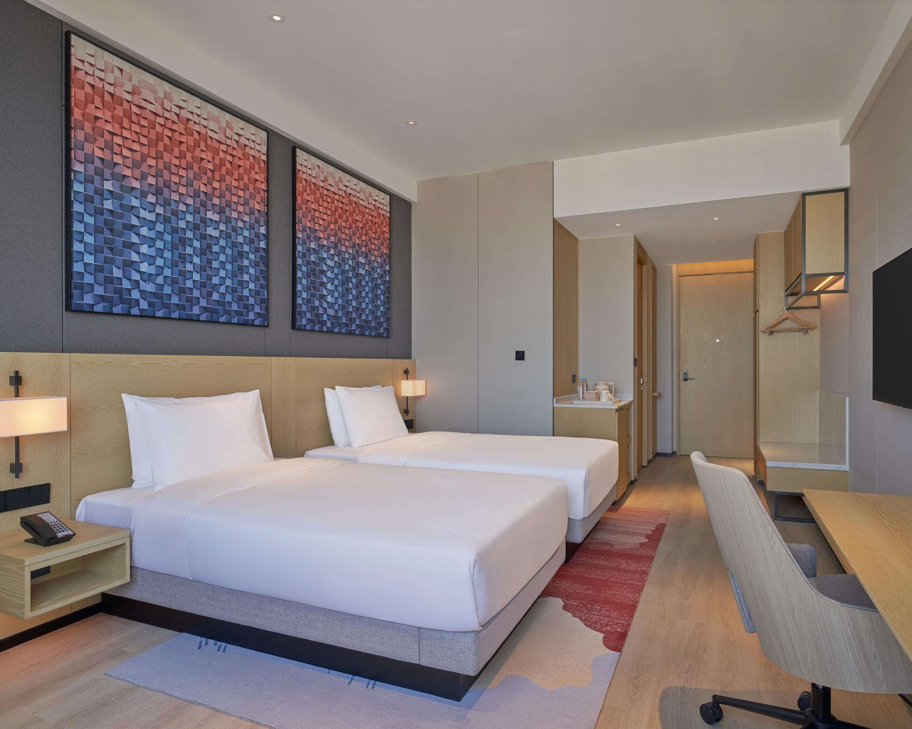 Quarto de hotel com duas camas, uma escrivaninha, uma cadeira, telefone na mesa de cabeceira e obras de arte na parede.