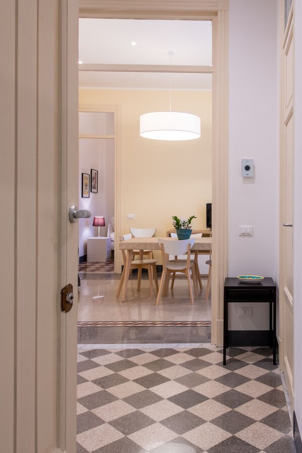 Um corredor com piso quadriculado leva a uma sala de jantar com mesa e cadeiras, um lustre e uma pequena mesa com uma planta.