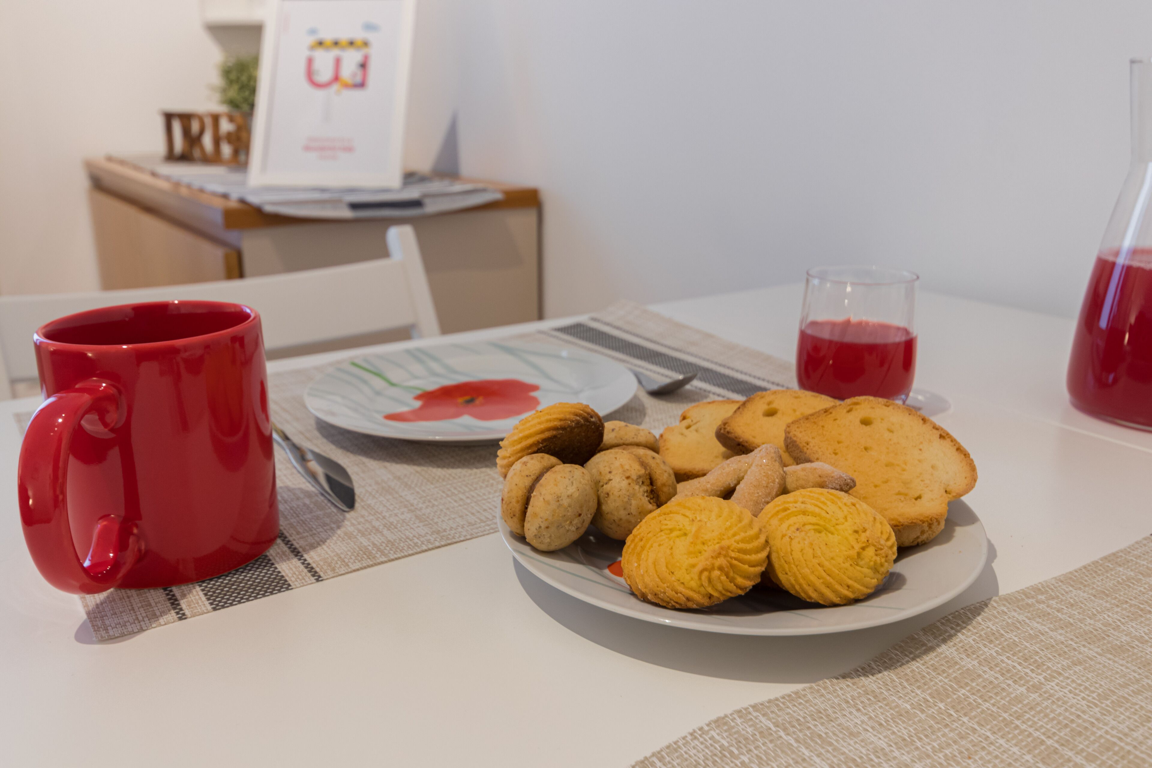 Uma mesa com um prato de biscoitos, uma caneca vermelha e um copo de bebida vermelha.