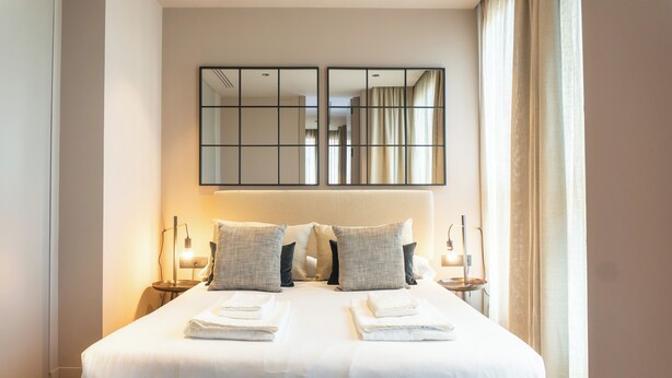 Uma cama bem arrumada com lençóis brancos e duas almofadas, uma mesa de cabeceira com abajur, e um espelho grande acima da cama.