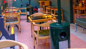 Um café com mesas e cadeiras de madeira, um cesto de lixo verde e luminárias fixadas na parede.