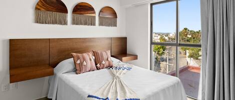 Quarto de hotel com uma cama grande, cabeceira de madeira, vista da cidade através de uma janela ampla e uma varanda com vista para a rua.