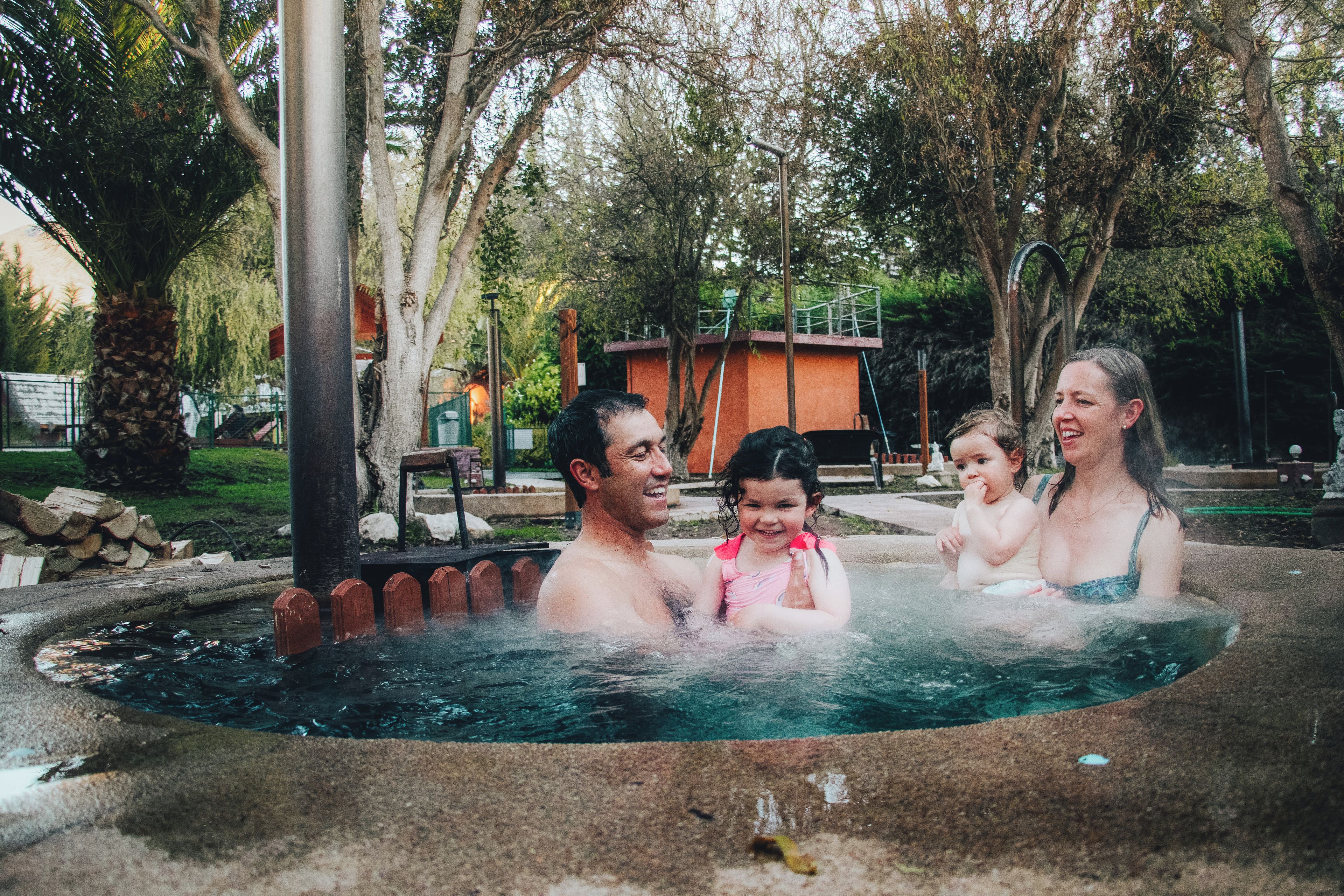 Uma família aproveitando uma banheira de hidromassagem em um parque com árvores e um edifício ao fundo.