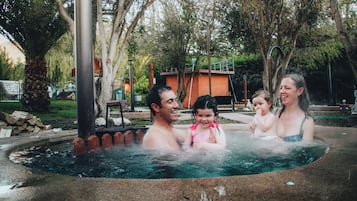 Uma família aproveitando uma banheira de hidromassagem em um parque com árvores e um edifício ao fundo.