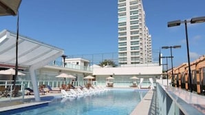 Uma piscina com espreguiçadeiras e guarda-sóis, cercada por edifícios modernos.