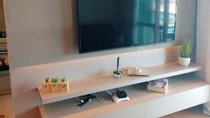 Uma sala de estar moderna com uma grande televisão de tela plana fixada em uma parede cinzenta. Há prateleiras abaixo da TV com objetos decorativos e uma pequena planta.