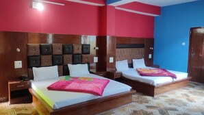 Quarto de hotel com duas camas, cada uma com cabeceira de madeira e roupa de cama branca. O quarto tem paredes vermelhas e azuis, e o chão possui um design de azulejos estampados.
