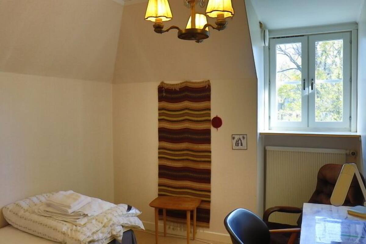 Quarto com uma cama, uma cadeira, uma escrivaninha com papéis, um radiador, uma janela com vista para árvores e uma tapeçaria pendurada.