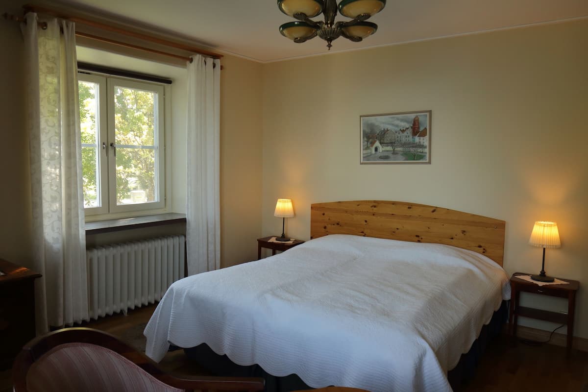 Quarto com cama grande, duas luminárias de cabeceira, painel de cabeceira de madeira, radiador, janela com cortinas e um quadro na parede.