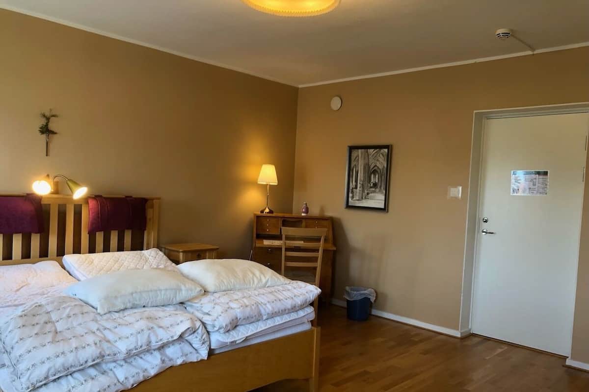 Quarto com cama de madeira, mesa de cabeceira, escrivaninha, cadeira, luminária, quadro emoldurado, porta e lixeira.