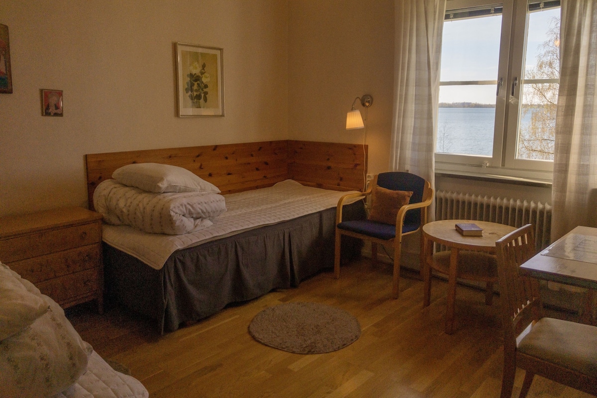 Um quarto acolhedor com uma cama de madeira, uma cadeira, uma mesinha e vista para a água pela janela.
