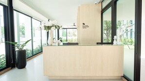 Uma receção com acabamento em madeira, bancada de vidro e placa com a inscrição "Pillows Boutique Hotel". Há plantas em vasos e vista para o exterior através de grandes janelas.