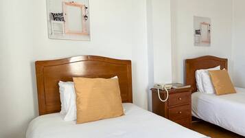 Duas camas de solteiro com cabeceiras de madeira, roupa de cama branca e uma pequena mesa de cabeceira com abajur.