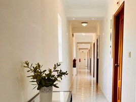 Um corredor comprido e estreito, paredes brancas e chão ladrilhado. Há uma planta em vaso à esquerda e uma porta de madeira à direita.
