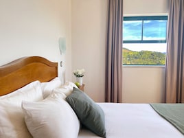 Uma cama bem arrumada com almofadas brancas e verdes, uma cabeceira de madeira e uma mesa de cabeceira com um vaso de flores e uma maçã.