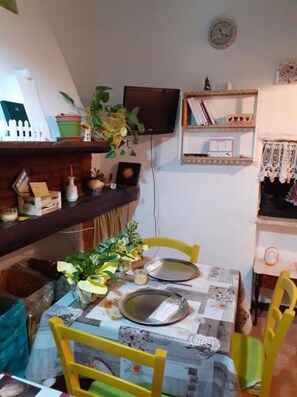Uma sala de jantar acolhedora com uma mesa posta para dois, rodeada por cadeiras amarelas. Uma televisão está montada na parede acima da mesa, e uma estante é visível à direita.