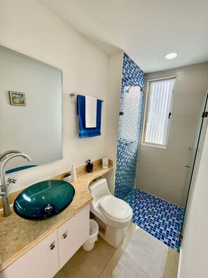 Casa de banho com chuveiro em azulejos azuis, sanita branca, lavatório com cuba azul e espelho.