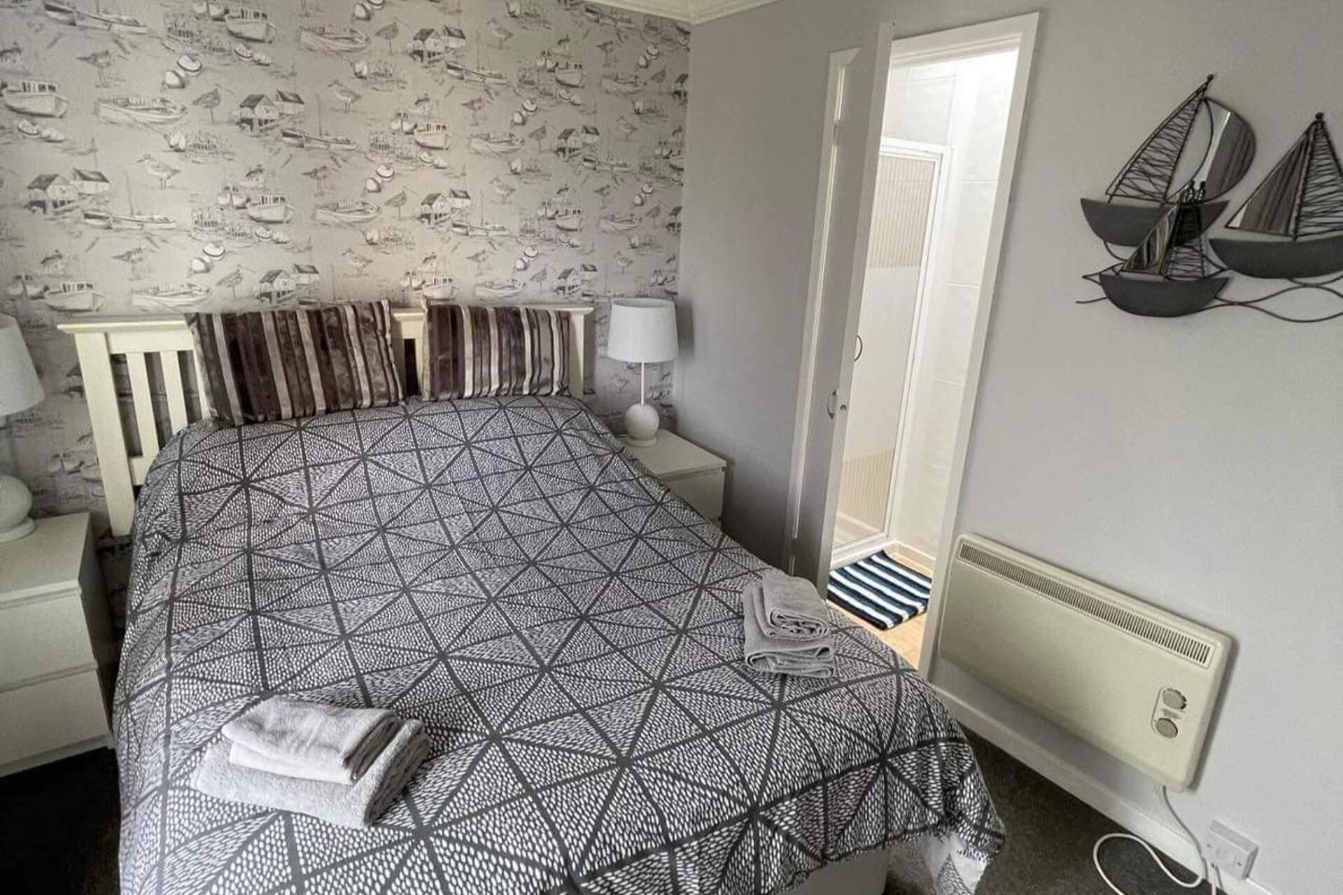 Quarto com colcha de padrão geométrico, mesa de cabeceira branca com luminária, radiador e parede com papel de parede de tema náutico.