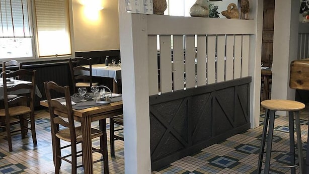 Interior de restaurante com mesas e cadeiras de madeira, área de bar com banquetas e parede decorativa com padrão geométrico.