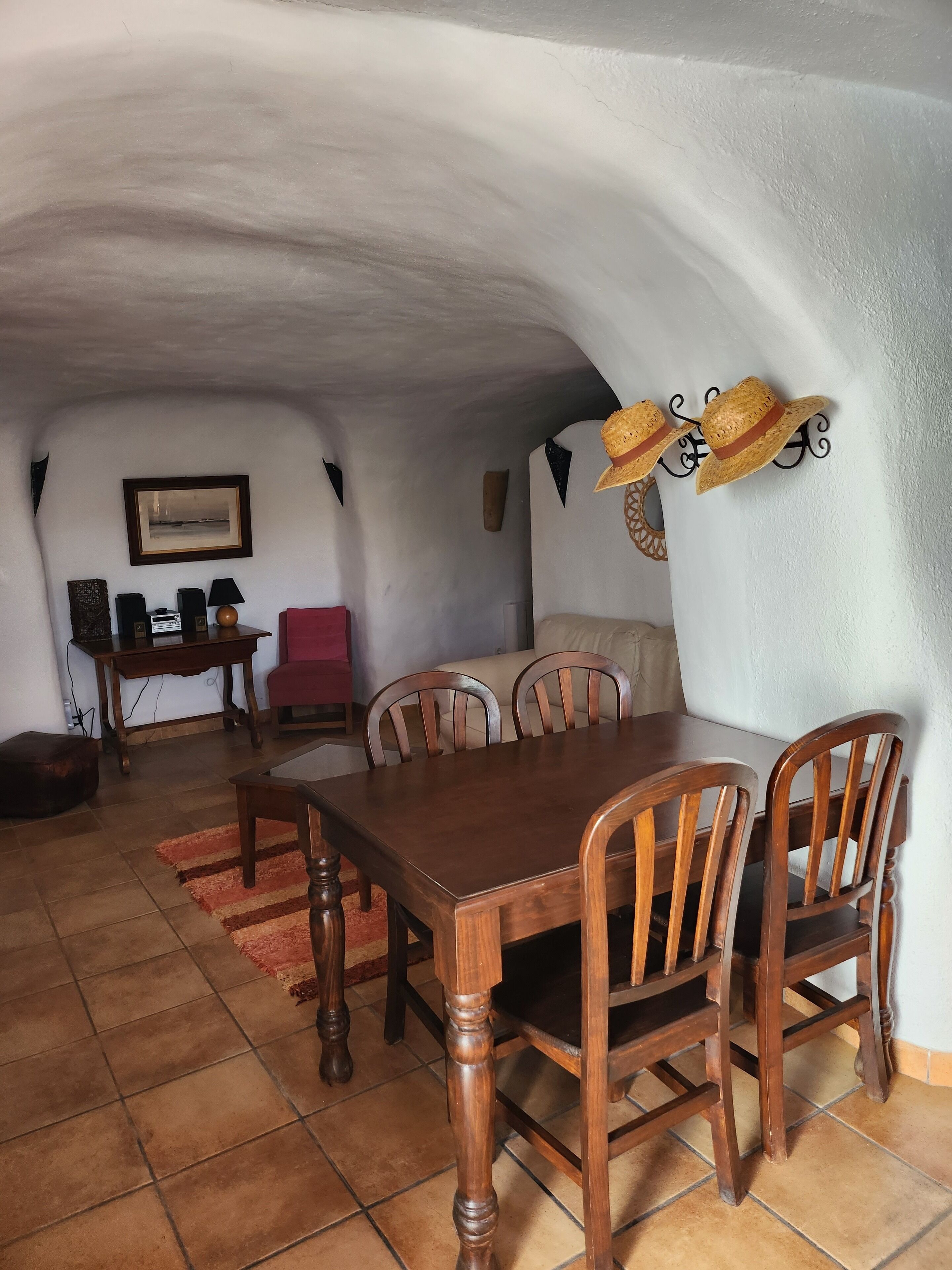 Uma área de refeições com mesa e cadeiras de madeira, uma cadeira vermelha, uma mesinha com rádio e luminária, e um quadro na parede.