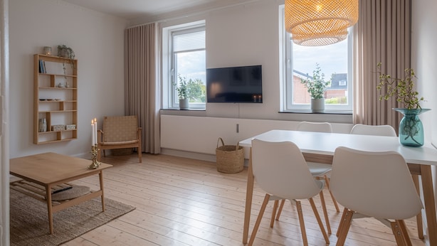 Uma sala de estar moderna com uma TV na parede, uma mesa de jantar com cadeiras, uma estante e uma janela grande com cortinas.