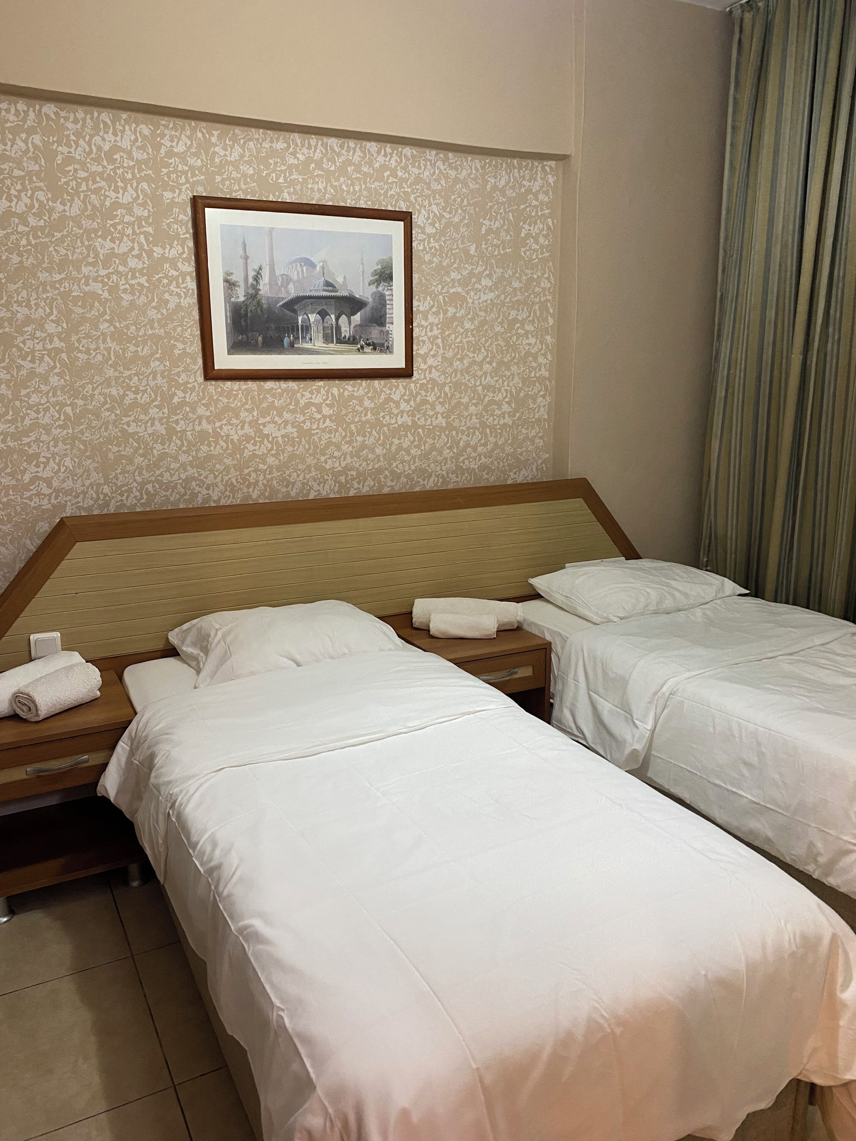 Quarto de hotel com duas camas, cabeceira de madeira, quadro emoldurado na parede e cortina listrada.