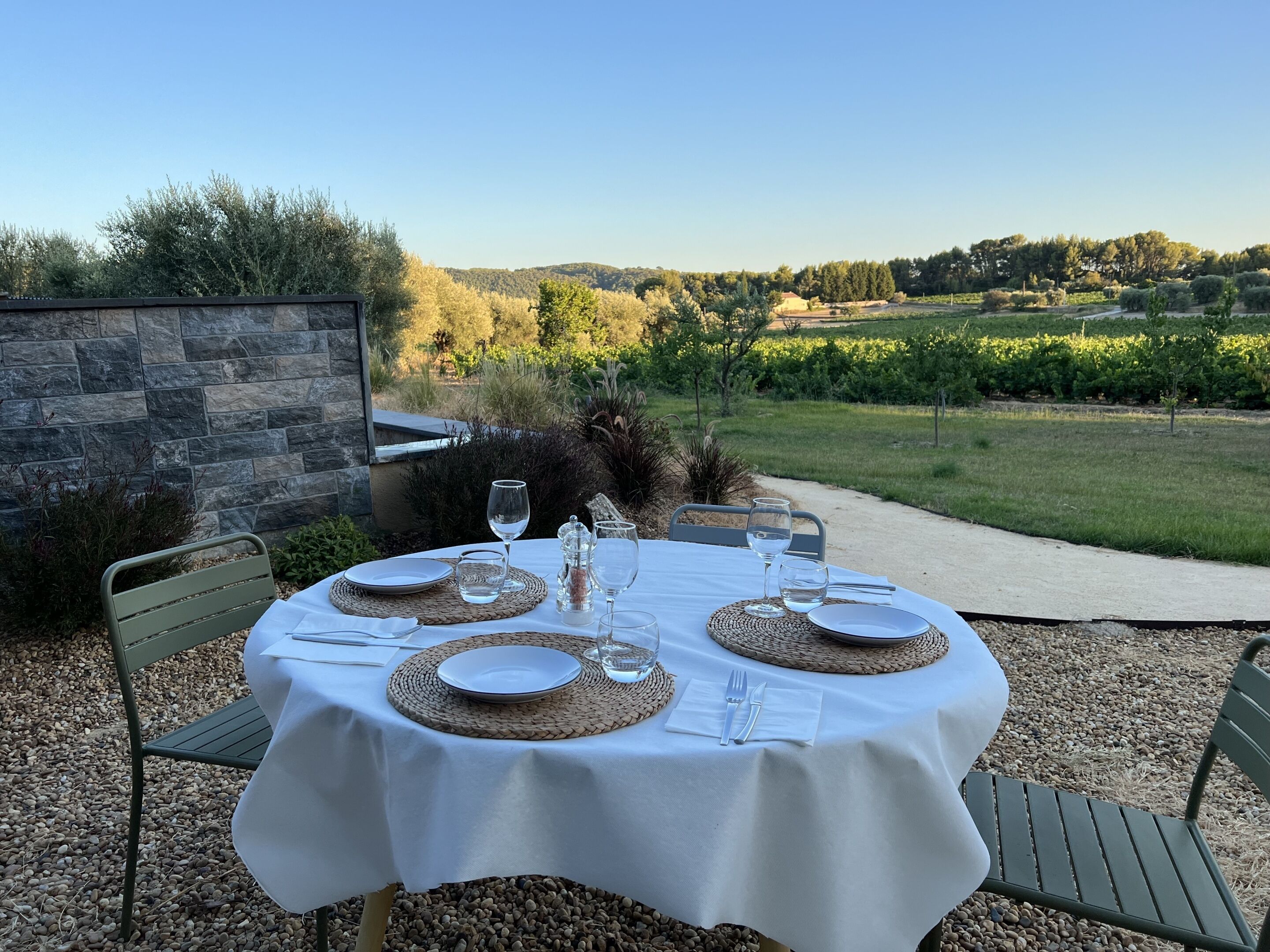 Mesa de jantar ao ar livre posta com pratos, copos e talheres, cercada por um muro de pedra e vista para um vinhedo.