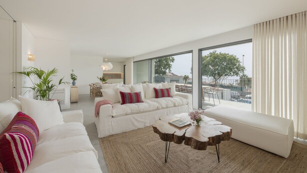 Uma sala de estar moderna com um sofá branco, uma mesa de centro de madeira e uma grande janela com vista para o exterior.