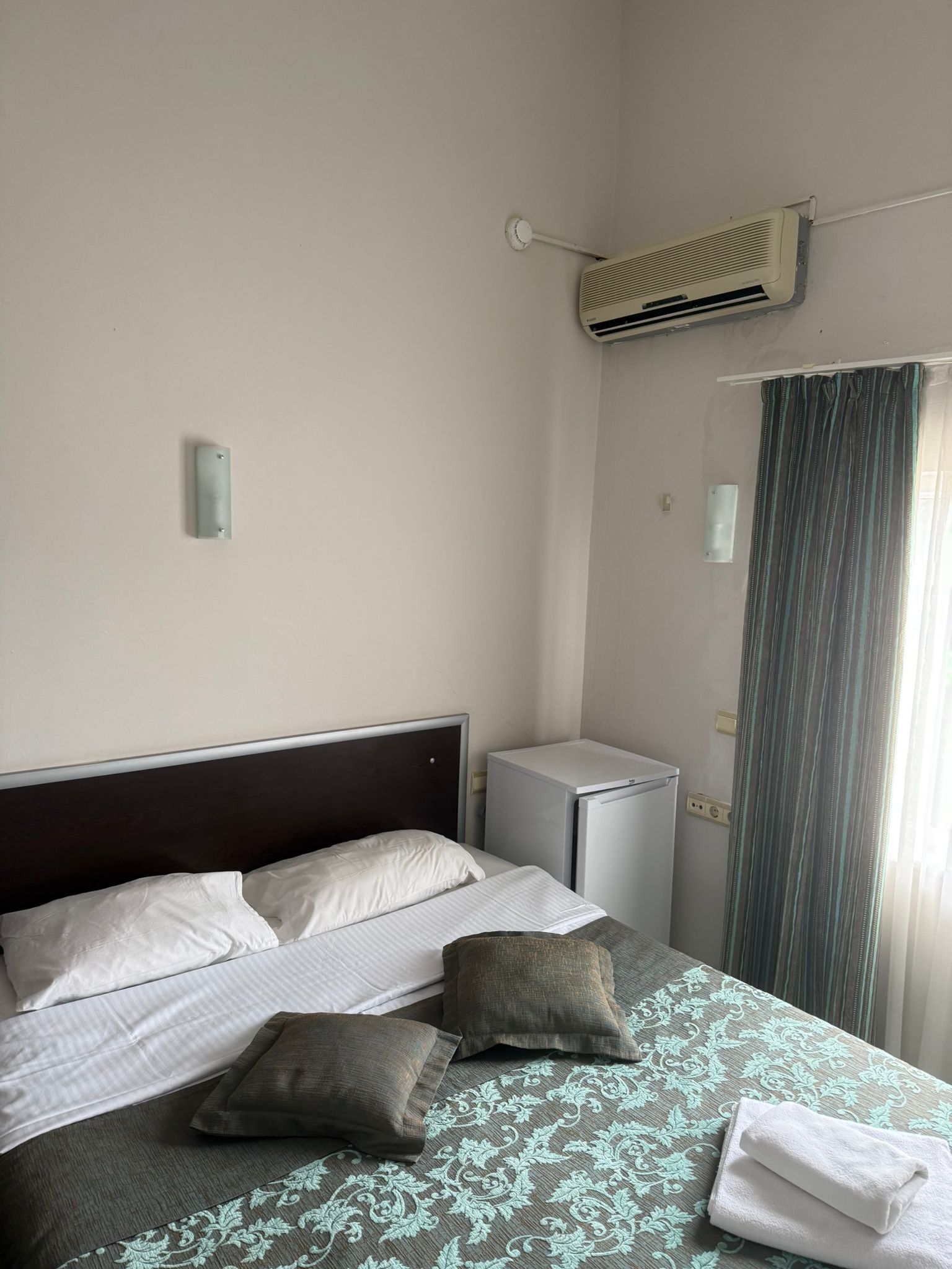 Quarto de hotel com cama, mesa de cabeceira, pequena geladeira, ar condicionado e janela com cortinas.