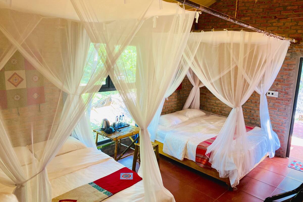Quarto com duas camas cobertas por dossel branco, uma mesa de madeira com toalha vermelha e parede de tijolos.