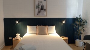 Quarto de hotel bem arrumado com uma cama grande, duas mesas de cabeceira, uma mesinha, uma cadeira e um vaso com planta.